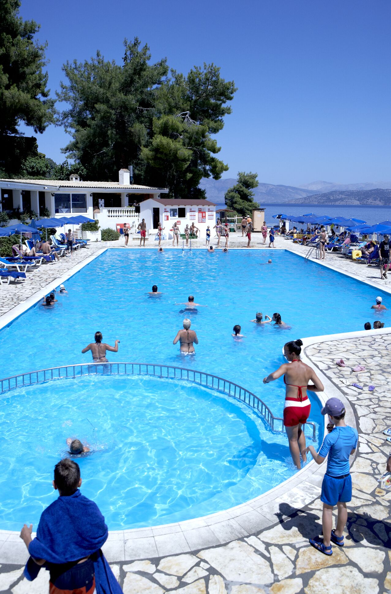 Piscina com pessoas na água e no deck, rodeada por guarda-sóis azuis e espreguiçadeiras.