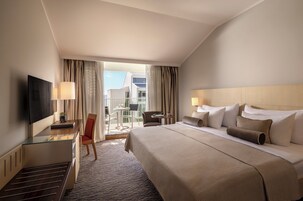 Quarto de hotel com cama, escrivaninha, televisão, cadeira e varanda com cortinas.