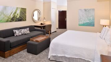 Quarto de hotel com uma cama grande, um sofá, uma escrivaninha e uma cadeira. Há duas pinturas nas paredes e uma porta ao fundo.