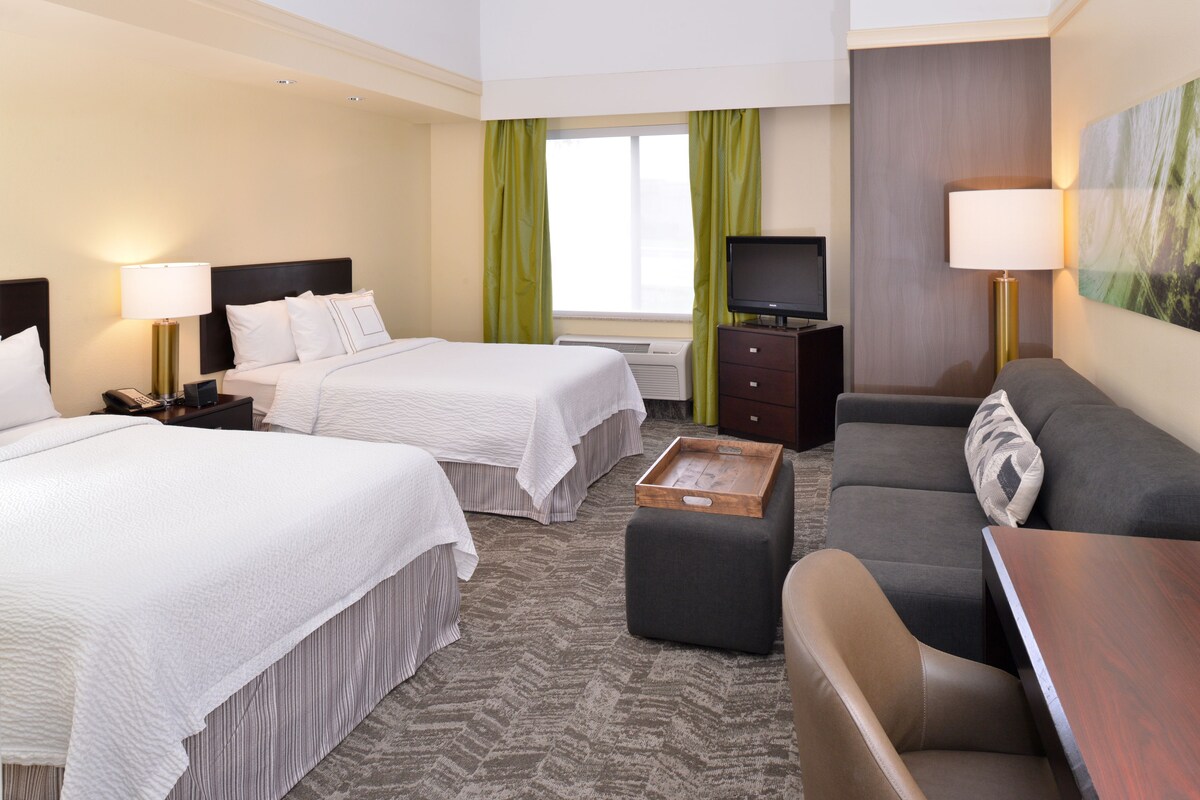 Quarto de hotel com duas camas, um sofá, uma TV e uma escrivaninha.