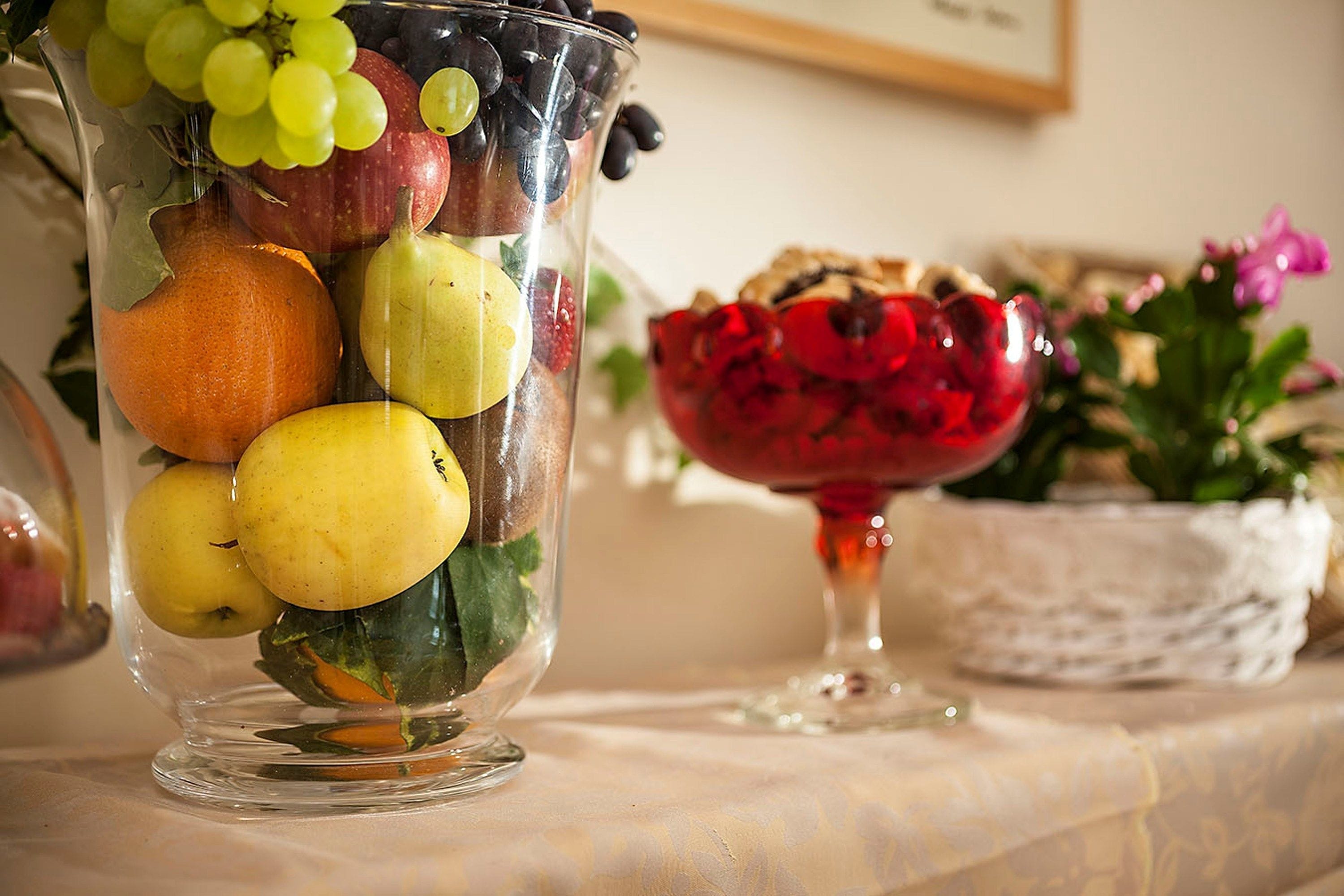 Um vaso de vidro transparente cheio de diversas frutas, como maçãs, uvas e laranjas, colocado sobre uma mesa com toalha de padrão decorativo.