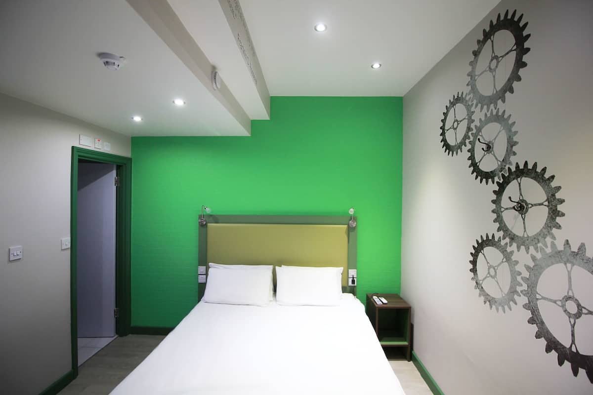 Quarto de hotel com parede verde, cama com lençóis brancos e decoração de engrenagens.