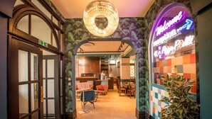 Um saguão de hotel com balcão de recepção, área de estar e um letreiro de neon indicando um bar e restaurante.