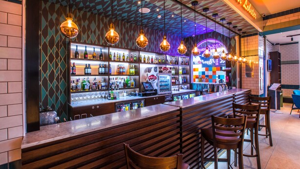 Uma área de bar com balcão, prateleiras cheias de garrafas, luminárias pendentes e banquetas.