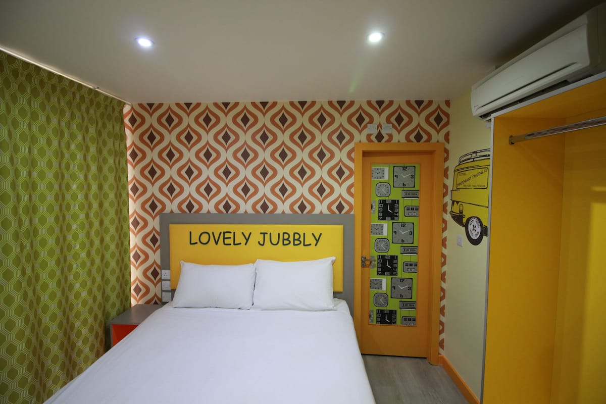 Quarto de hotel com cama, cabeceira com a inscrição 'LOVELY JUBBLY', relógio de parede e porta com uma placa amarela.