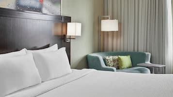Quarto de hotel com uma cama grande, uma poltrona verde, uma mesa pequena e uma luminária fixada na parede.