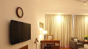 Quarto de hotel com televisão de tela plana, escrivaninha com abajur, cadeira e uma janela grande com cortinas.