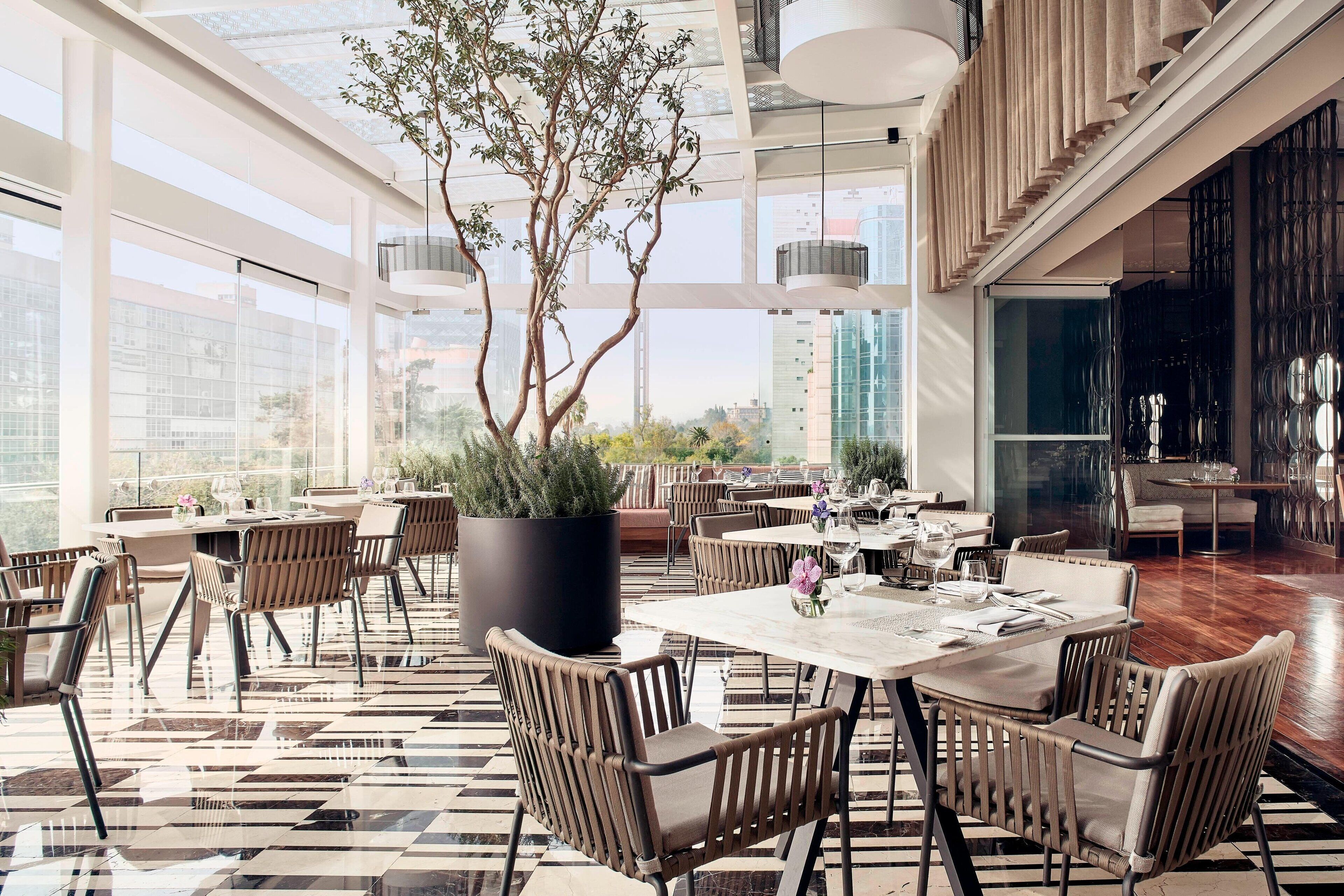 Restaurante moderno com pé-direito alto, grandes janelas e um vaso de plantas central. Mesas e cadeiras dispostas para refeições.