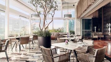 Restaurante moderno com pé-direito alto, grandes janelas e um vaso de plantas central. Mesas e cadeiras dispostas para refeições.