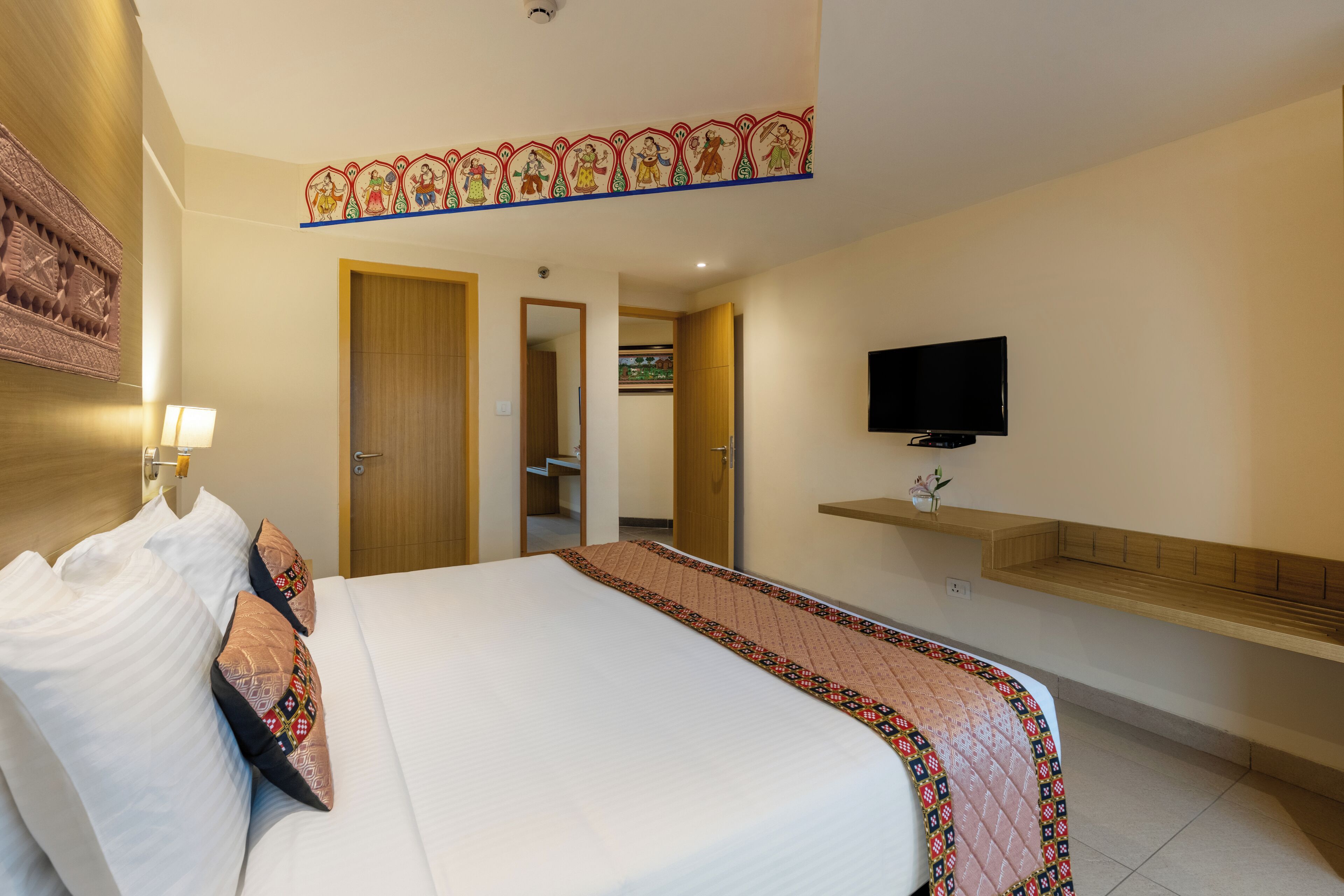 Quarto de hotel com uma cama grande, televisão, prateleira de madeira e teto decorativo.