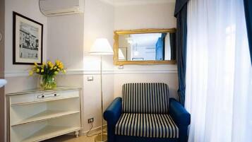 Quarto com poltrona azul, mesa consola branca com um vaso de flores amarelas, abajur e espelho na parede.