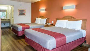 Quarto de hotel com duas camas, cabeceira de madeira, mesa de cabeceira e luminária fixada na parede.