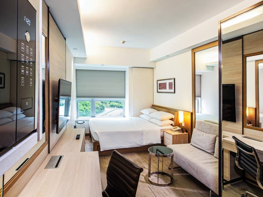 Quarto de hotel com cama, escrivaninha, cadeira, TV, espelho e janela com vista para o verde.