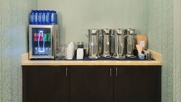 Uma área de bar com geladeira abastecida com bebidas, dispensadores de café e uma garrafa de xarope adocicado.