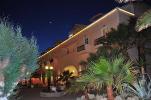 Um hotel com fachada branca, várias varandas e uma área de entrada bem iluminada, com palmeiras e uma fonte.