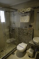 Casa de banho com chuveiro envidraçado, sanita com etiqueta a indicar "esterilizado", lavatório e um porta-toalhas com toalhas dobradas.
