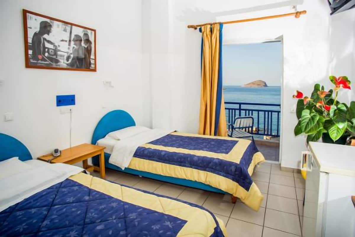 Quarto de hotel com duas camas, uma mesa pequena, uma cadeira, varanda com vista para o mar e um vaso de planta.