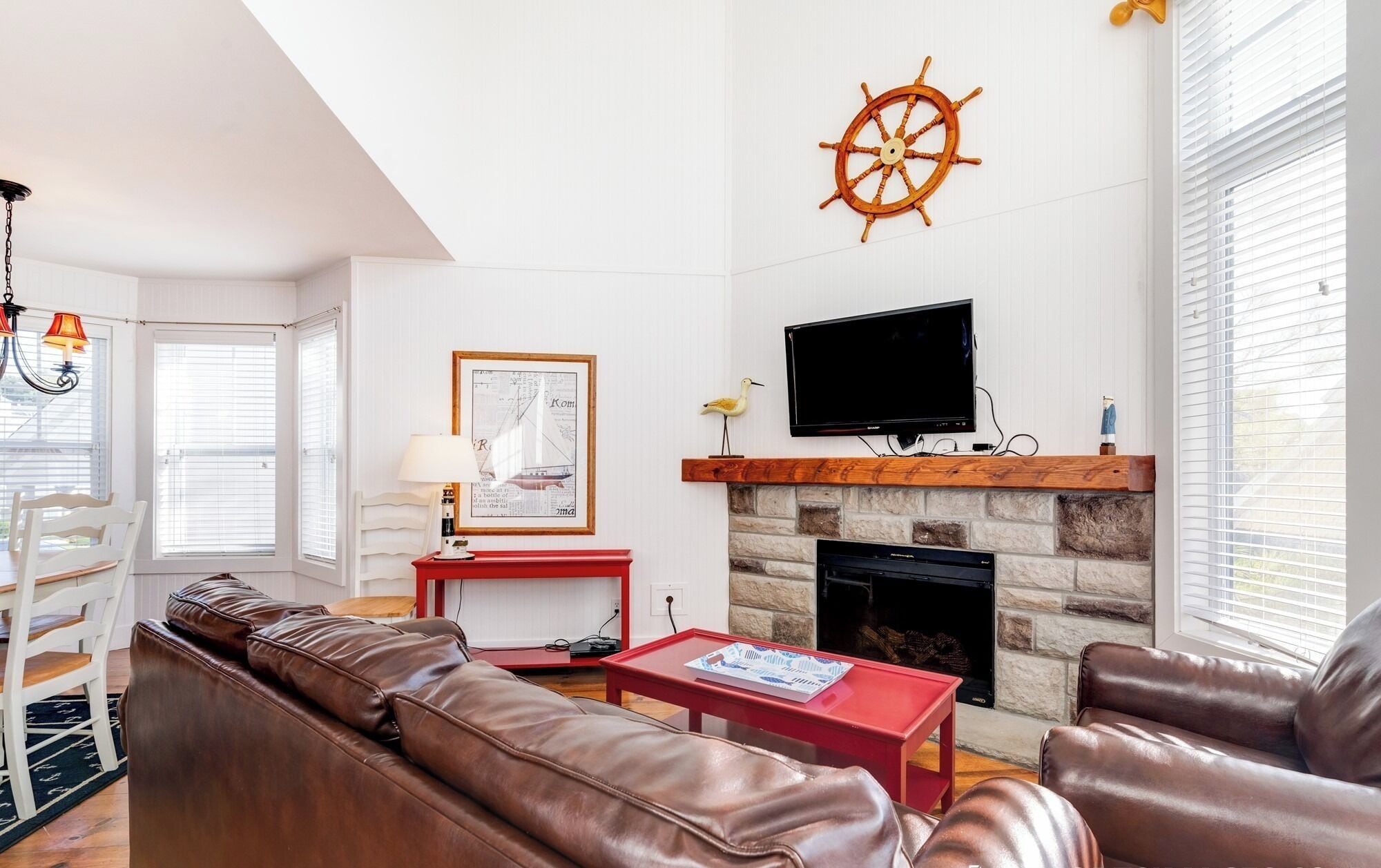 Uma sala de estar com sofá de couro, uma mesa vermelha, lareira, televisão montada e decoração de roda de navio.