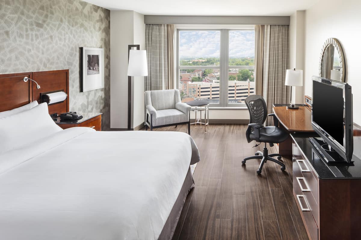 Quarto de hotel com uma cama grande, uma mesa com televisão, uma cadeira, uma luminária de mesa e vista para a cidade pela janela.