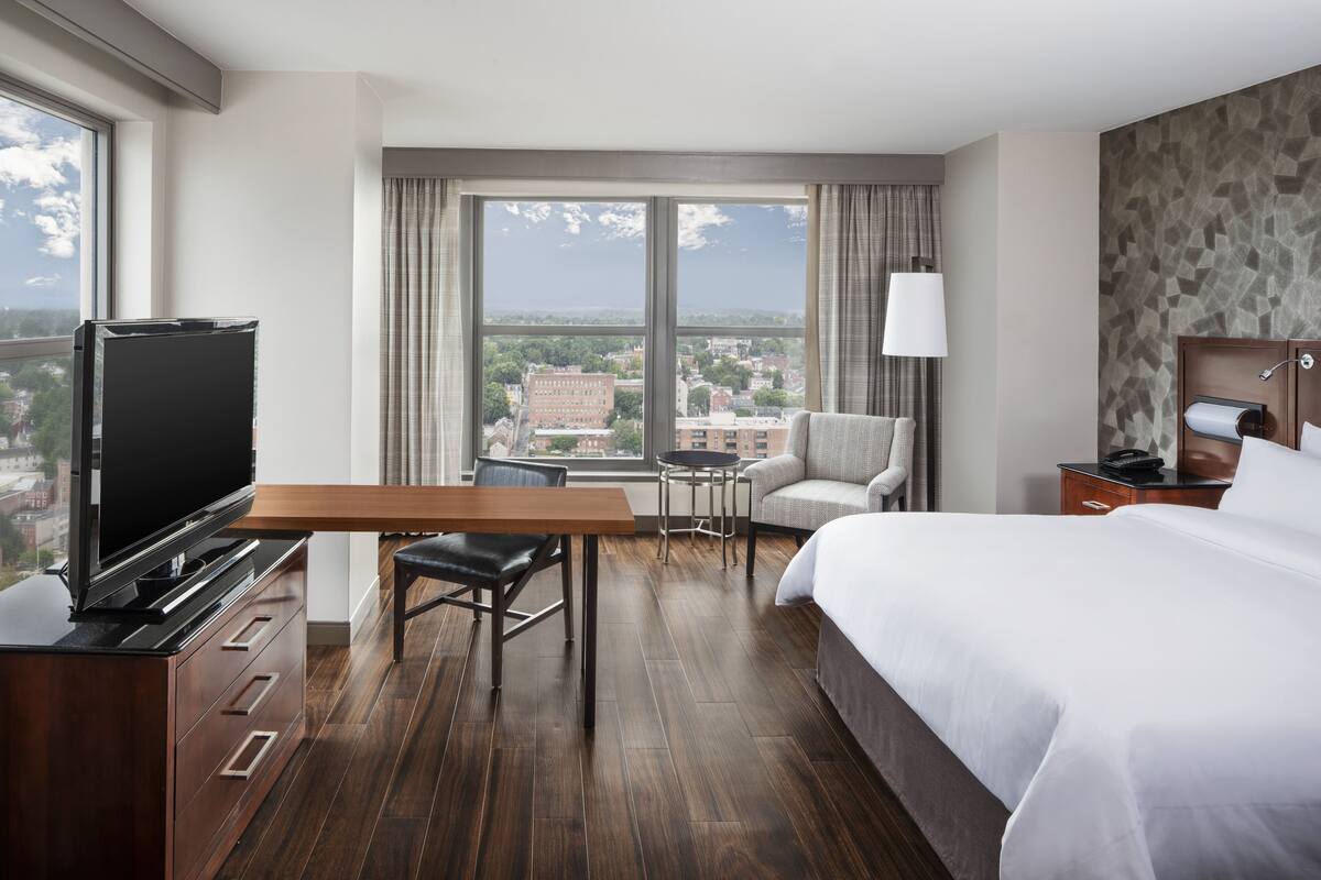 Quarto de hotel com uma cama grande, televisão em cômoda de madeira, mesinha com cadeira, sofá e vista da cidade por grandes janelas.