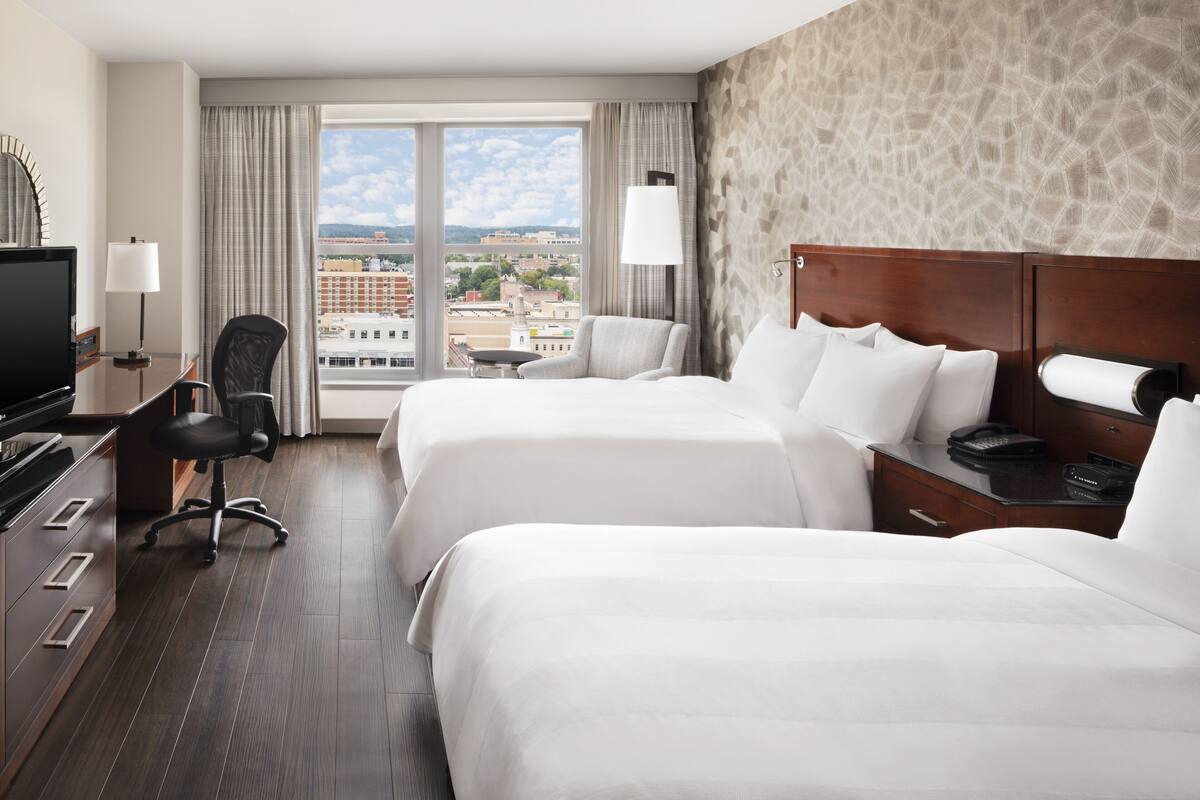 Quarto de hotel com duas camas, uma mesa de trabalho, uma televisão, uma cadeira e vista para a cidade.