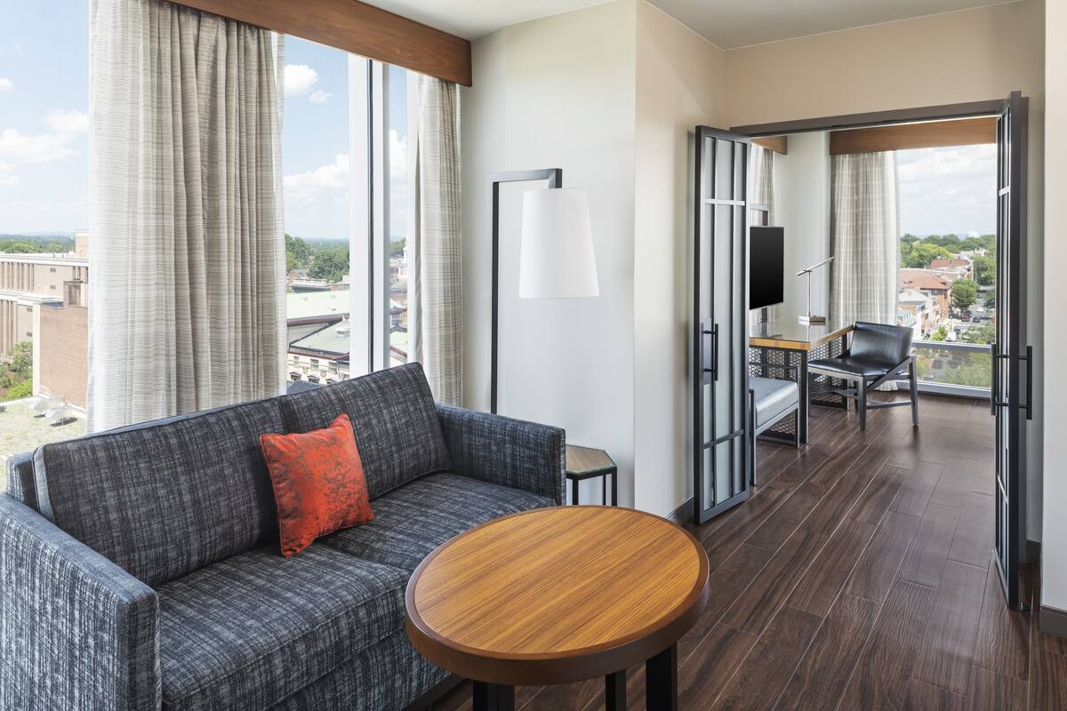 Quarto de hotel moderno com sofá cinzento, mesa redonda de madeira e varanda com vista para a cidade.