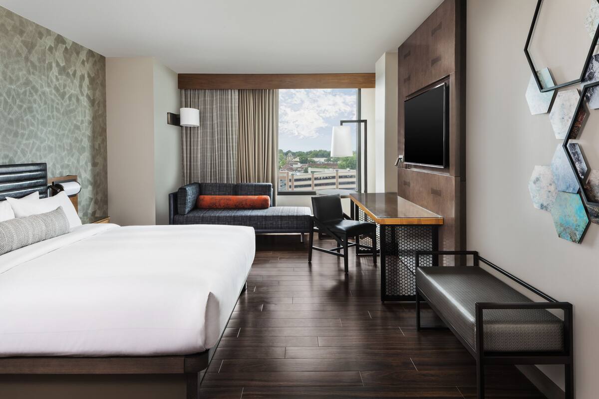 Quarto de hotel com uma cama grande, uma escrivaninha com cadeira, uma televisão e vista para a cidade.