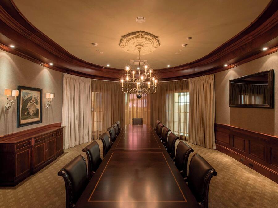 Uma sala de conferência com uma mesa comprida, cadeiras de couro, um candelabro e uma grande janela com cortinas.