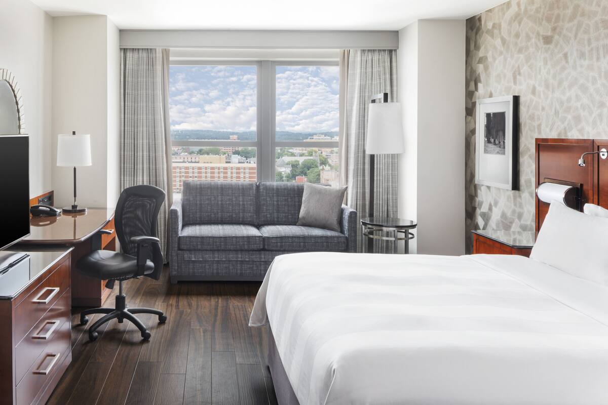 Quarto de hotel com uma cama grande, um sofá, uma escrivaninha com computador, uma cadeira, uma televisão e vista da cidade através de grandes janelas.