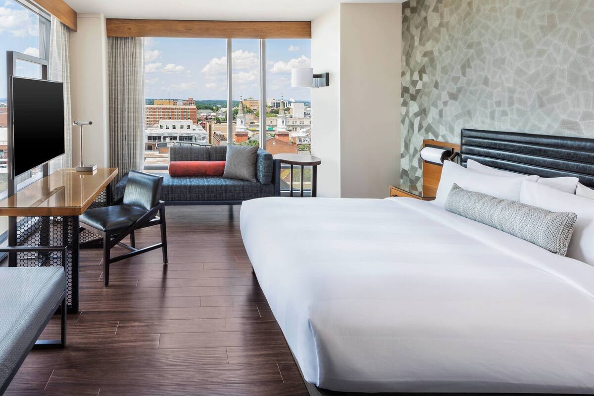 Quarto de hotel com uma cama grande, uma mesa com televisão de tela plana, um sofá e vista para a cidade.