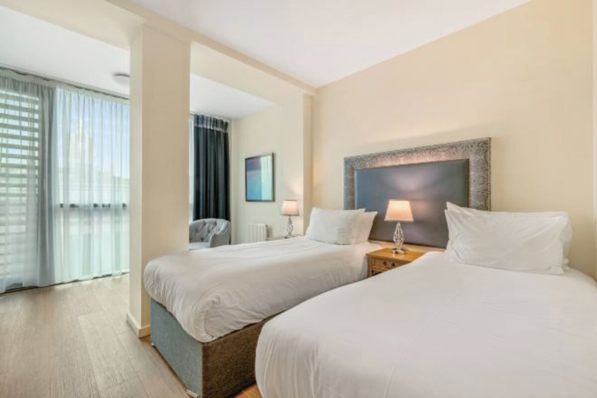 Quarto de hotel com duas camas, uma janela grande com cortinas, uma cadeira e um quadro na parede.