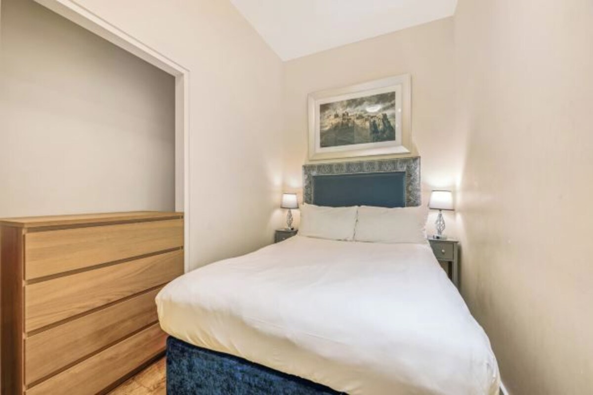 Quarto com cabeceira de madeira, colcha branca, duas luminárias de apoio e um quadro na parede.