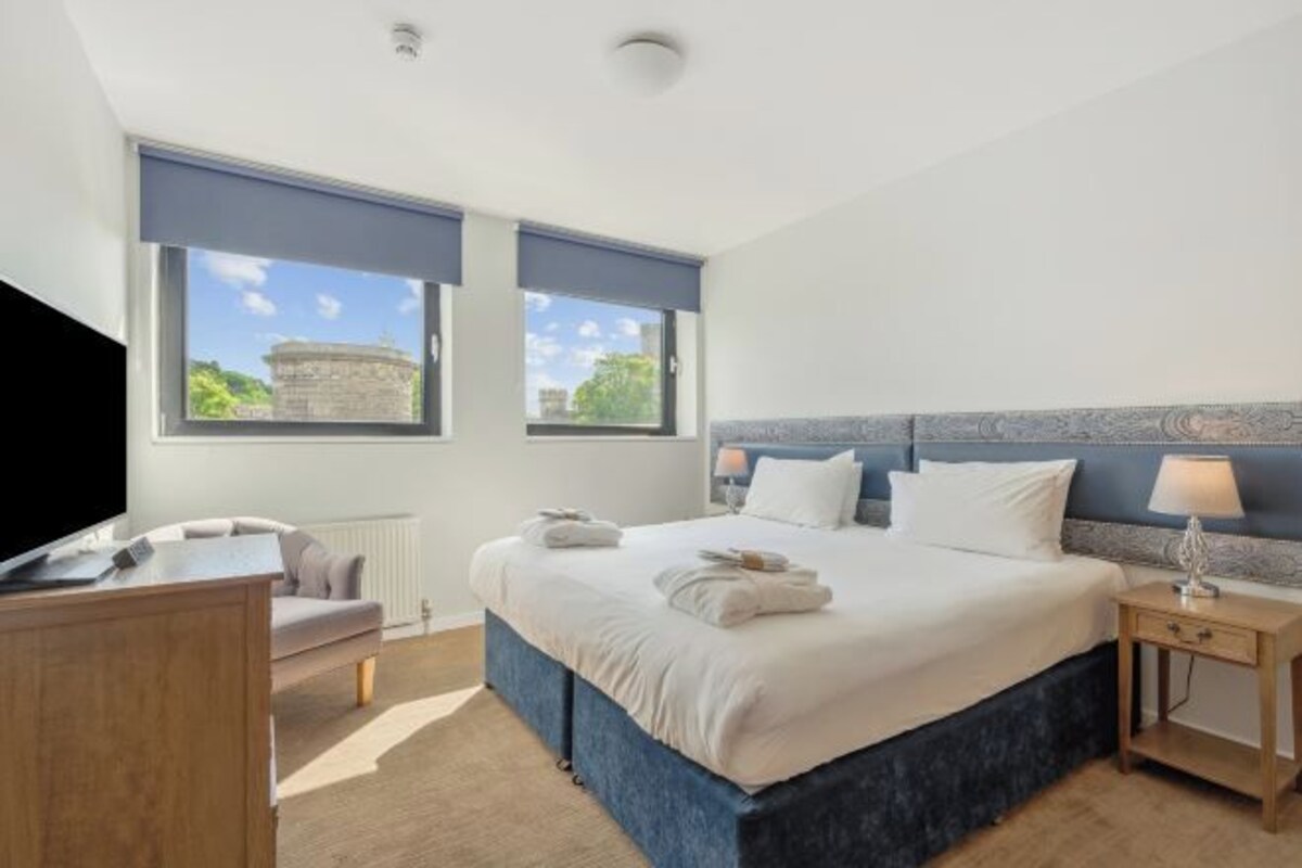 Quarto de hotel moderno com uma cama grande, uma escrivaninha e uma cadeira. Há duas janelas com persianas azuis, e vista para um edifício de pedra.