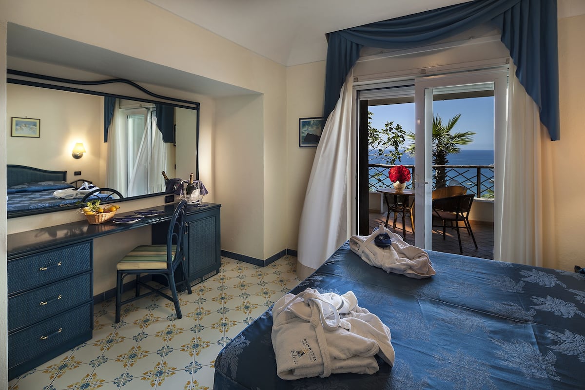 Quarto de hotel com cama, escrivaninha, cadeira, cômoda e varanda com vista para o mar.