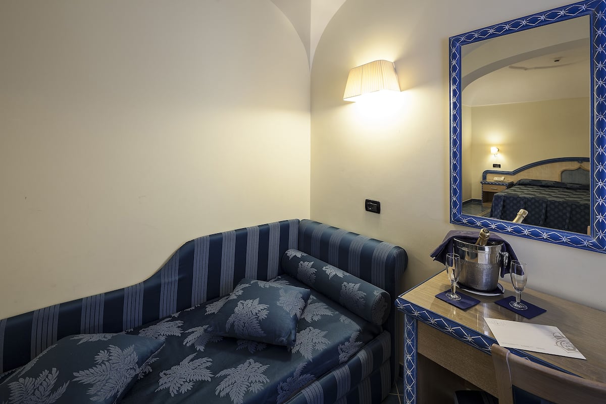 Quarto de hotel com sofá, mesa de cabeceira com luminária, espelho e minibar.