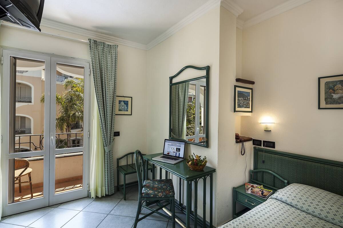 Um quarto com uma cama, uma escrivaninha com um laptop, uma cadeira, uma mesinha de apoio com um cesto, um telefone e uma janela com cortinas.