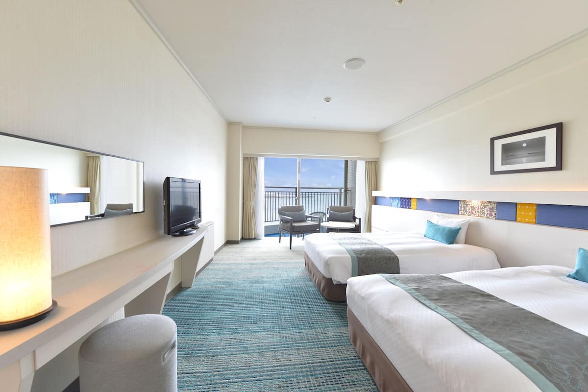 Quarto de hotel com duas camas, uma escrivaninha com luminária, televisão e varanda com vista.