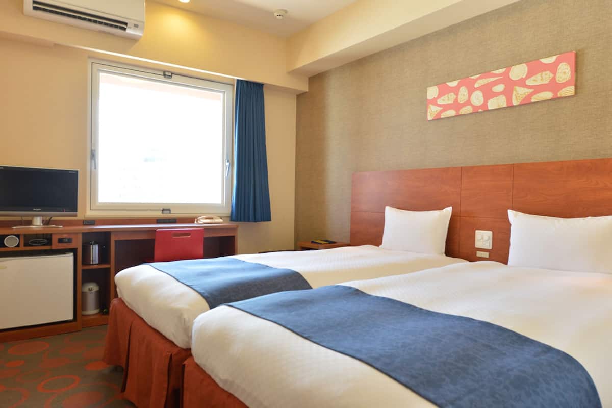 Quarto de hotel com duas camas, uma mesa de escritório com computador, televisão e janela com cortinas.