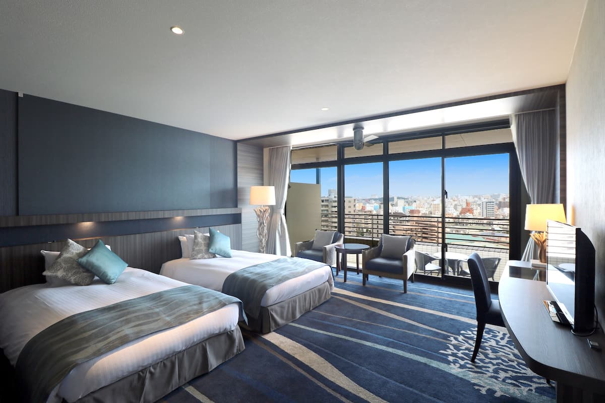 Quarto de hotel com duas camas, uma escrivaninha, uma cadeira e vista para a cidade.