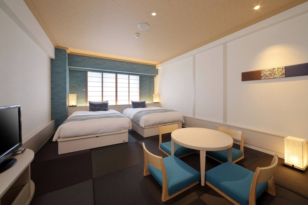 Quarto de hotel com duas camas, televisão, mesa redonda com cadeiras e janela com persianas.