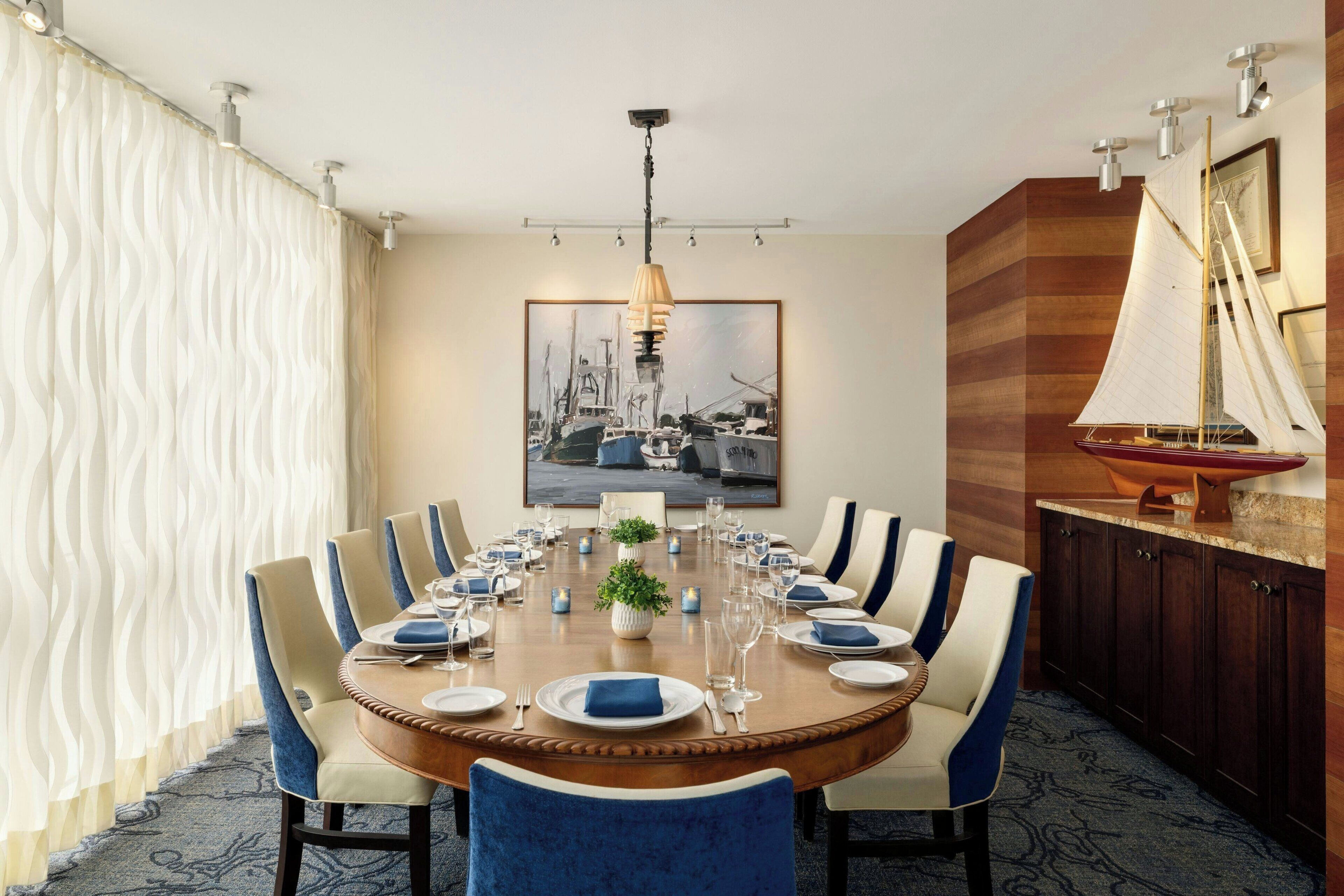 Uma sala de jantar formal com uma mesa redonda, cadeiras azuis e bege, e um quadro com tema náutico na parede.