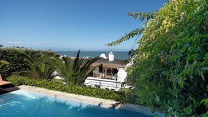 Piscina com vista para o mar, rodeada por vegetação e um edifício branco com telhado.