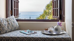 Assento junto à janela com vista para o oceano, um livro, óculos, um bule e uma xícara.
