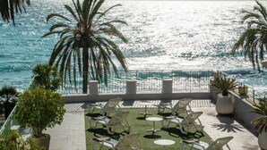Um terraço com cadeiras e mesas de descanso brancas, palmeiras e vista para o mar.