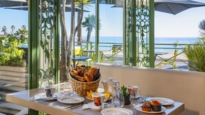Mesa de café da manhã com croissants, uma xícara de café e uma cesta de pão, com vista para uma praia com palmeiras.