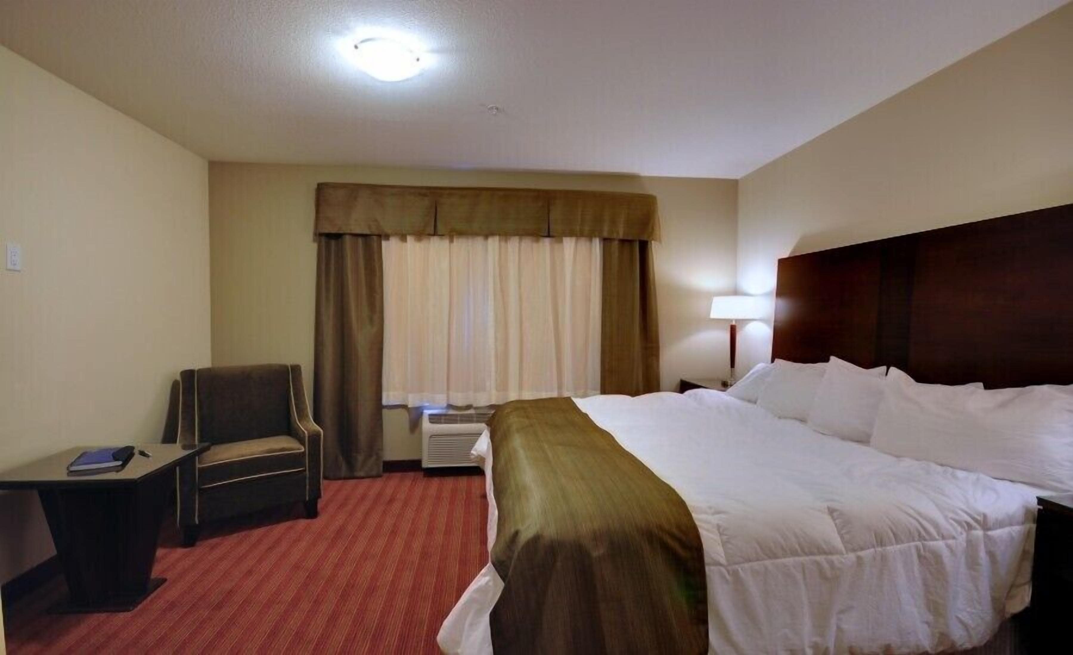 Quarto de hotel com cama, escrivaninha, cadeira, luminária e janela com cortinas.