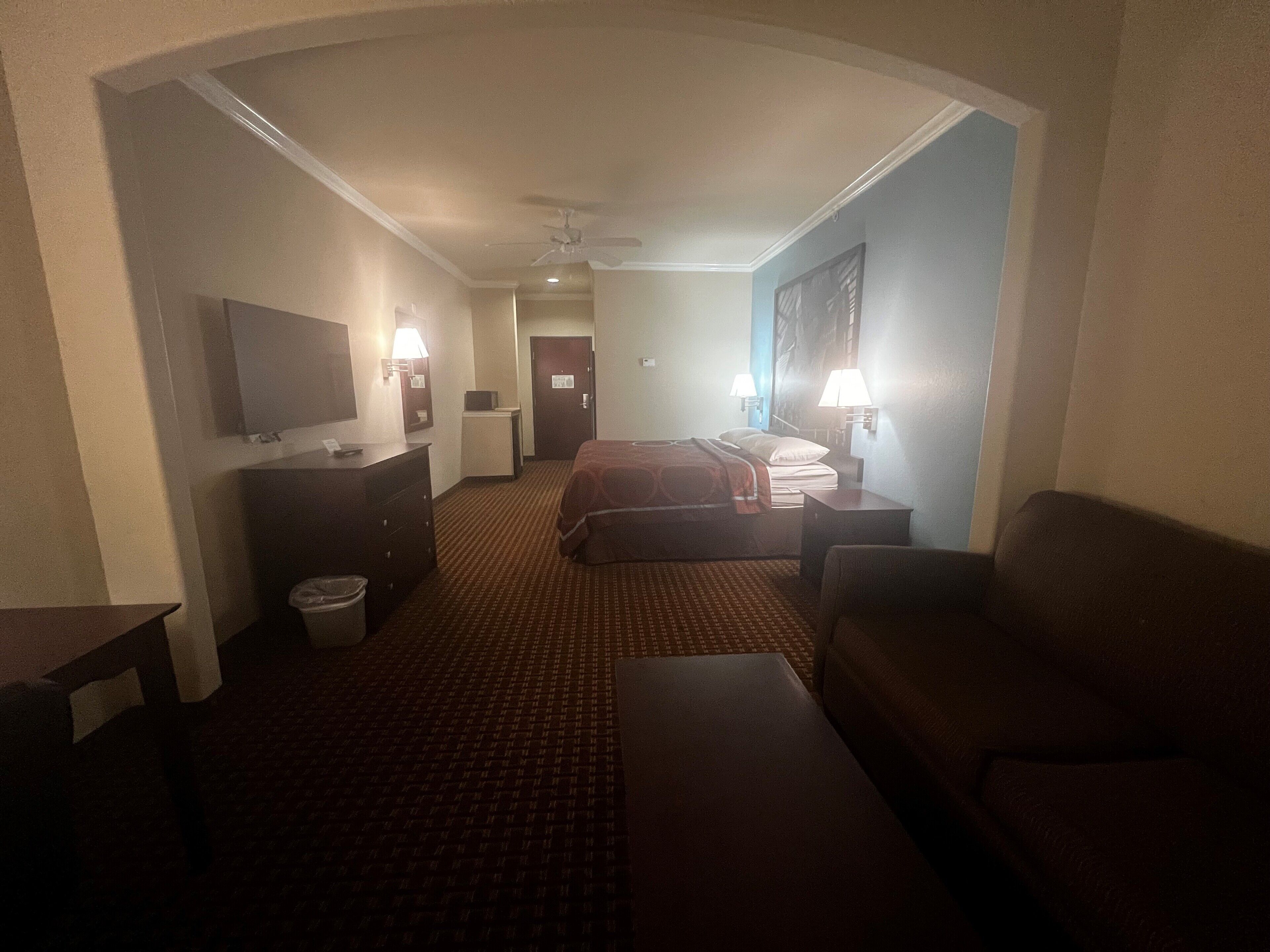 Quarto de hotel com cama, escrivaninha, cadeira, televisão e lixeira.