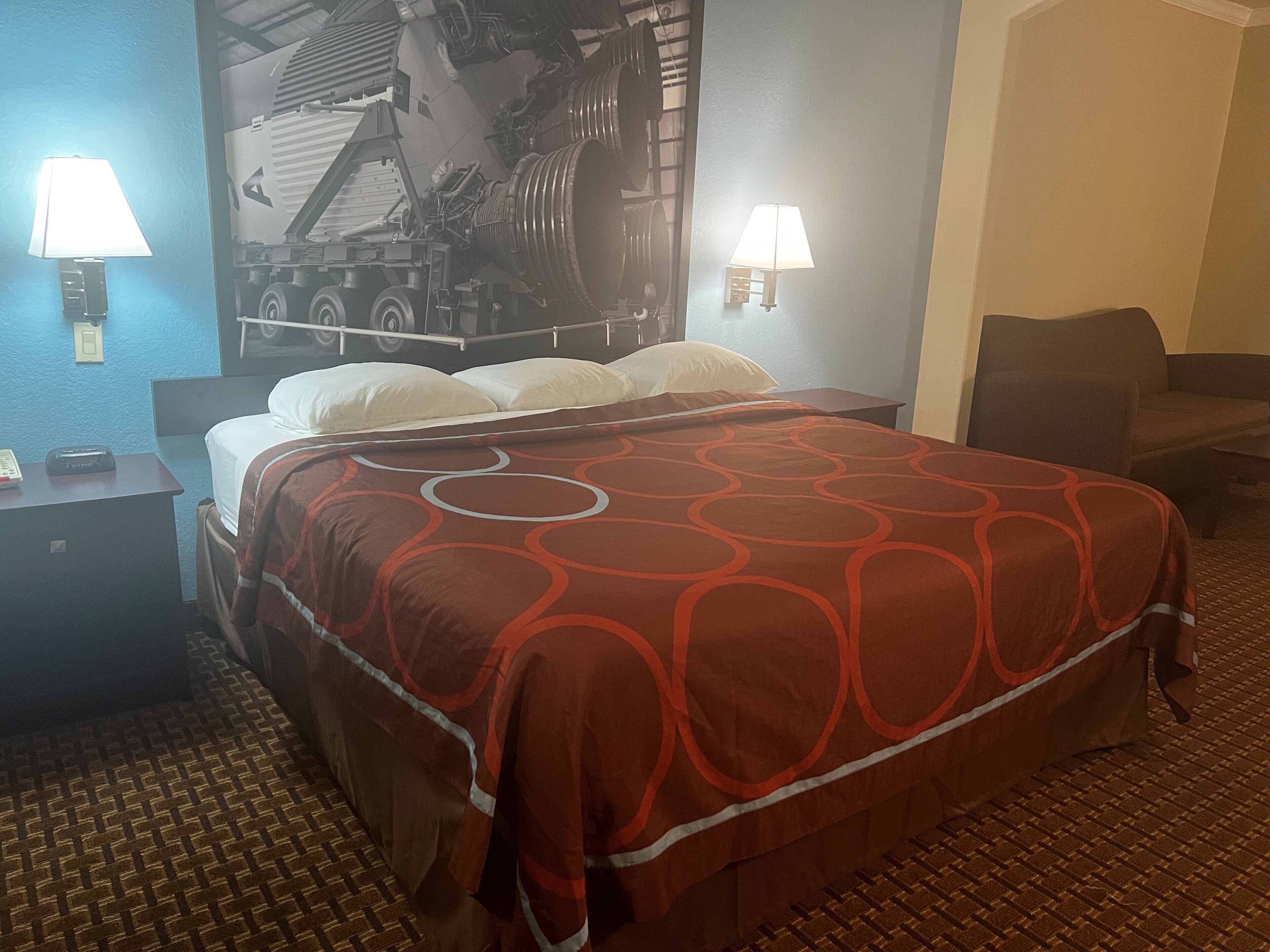 Quarto de hotel com uma cama, duas luminárias de cabeceira, uma mesa de cabeceira, uma TV fixada na parede e um pôster grande de um foguete.
