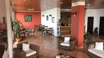 Um hall amplo com móveis de vime, área de bar com prateleiras cheias de garrafas e uma grande planta em vaso.
