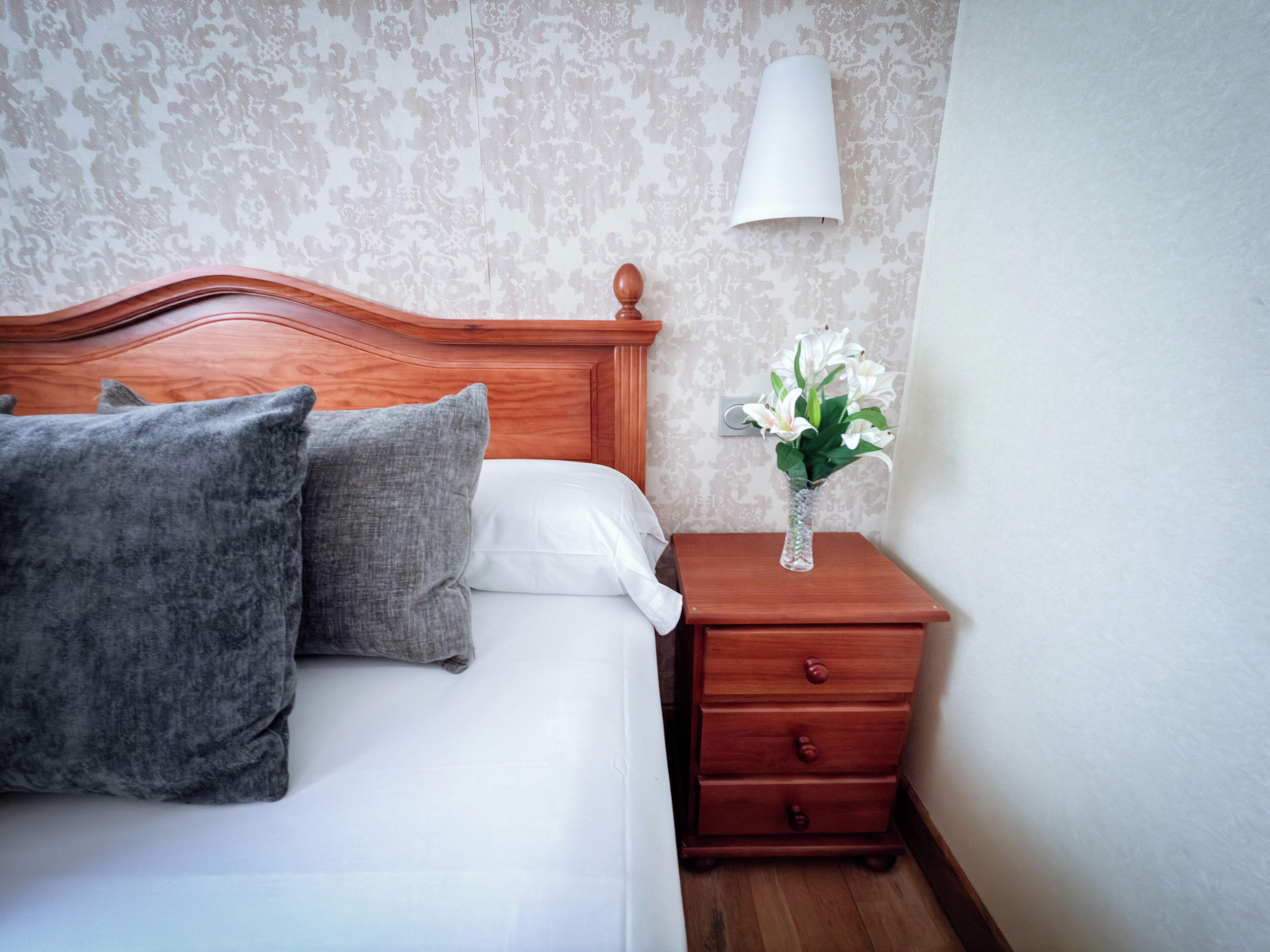 Uma cama bem arrumada com almofadas cinzas, uma mesa de cabeceira de madeira com um vaso de flores brancas e uma luminária fixada na parede.
